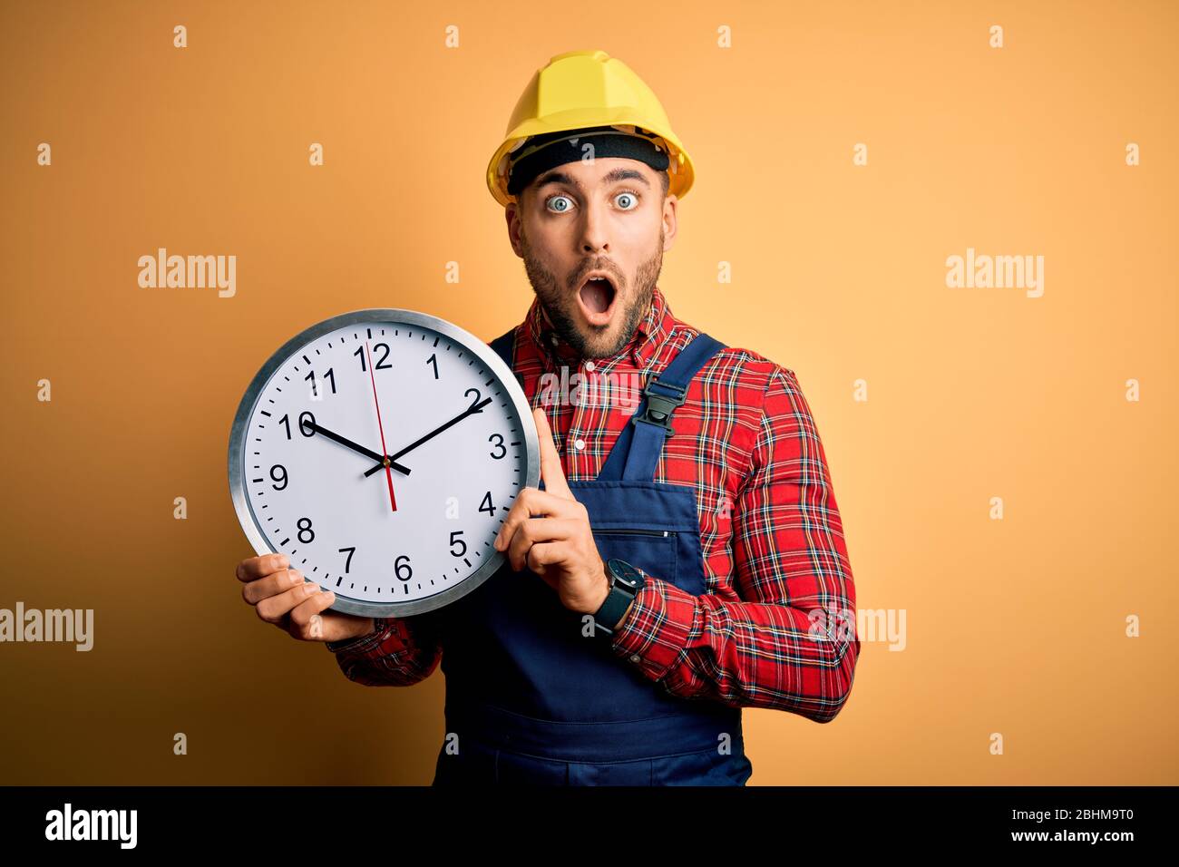 Young builder man wearing safety helmet holding big clock over yellow ...