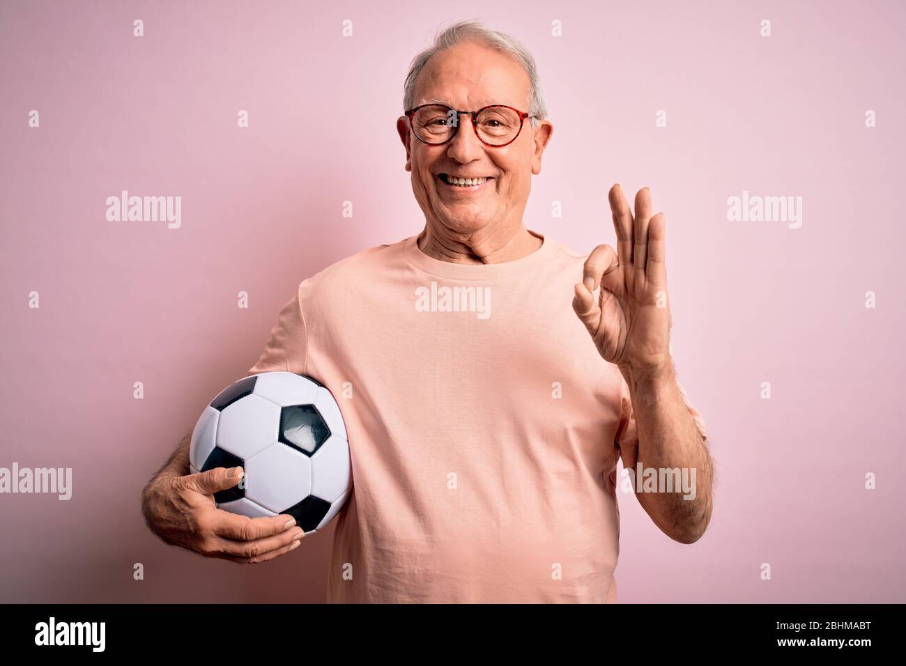 Grey haired senior football player man holding soccer ball over pink ...