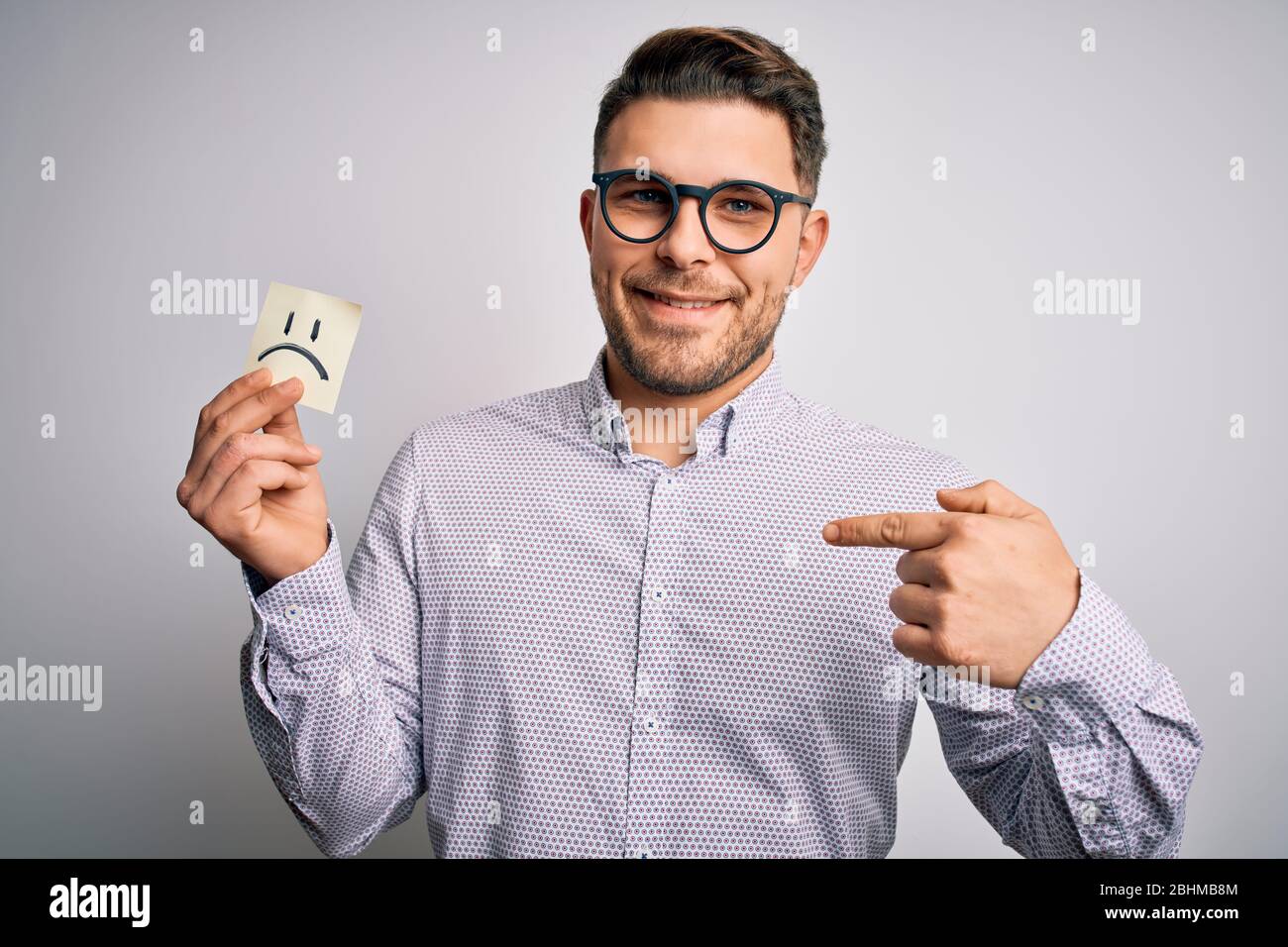 Young business man with blue eyes holding paper note with sad face as ...