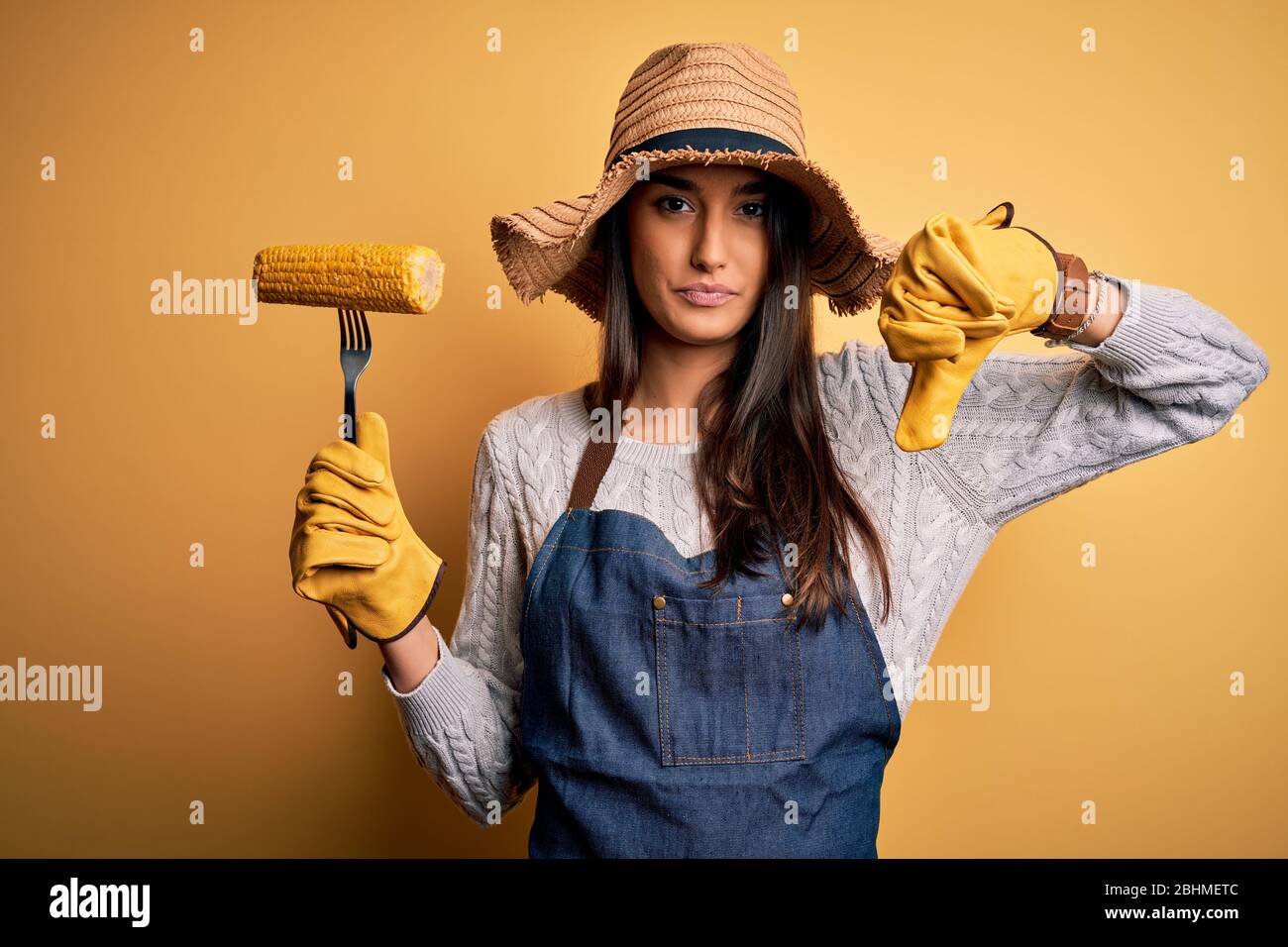 Young beautiful farmer woman wearing apron and hat holding fork with ...