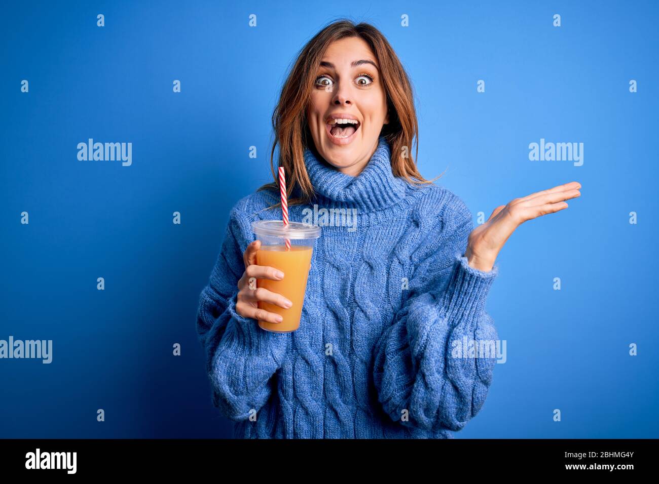 Young beautiful brunette woman drinking healthy orange juice over blue ...
