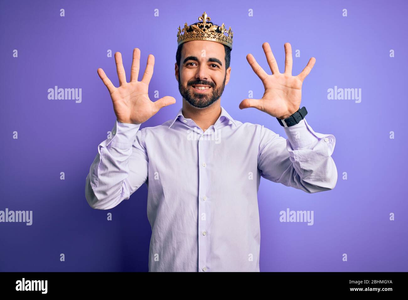 Young handsome man with beard wearing golden crown of king over purple ...