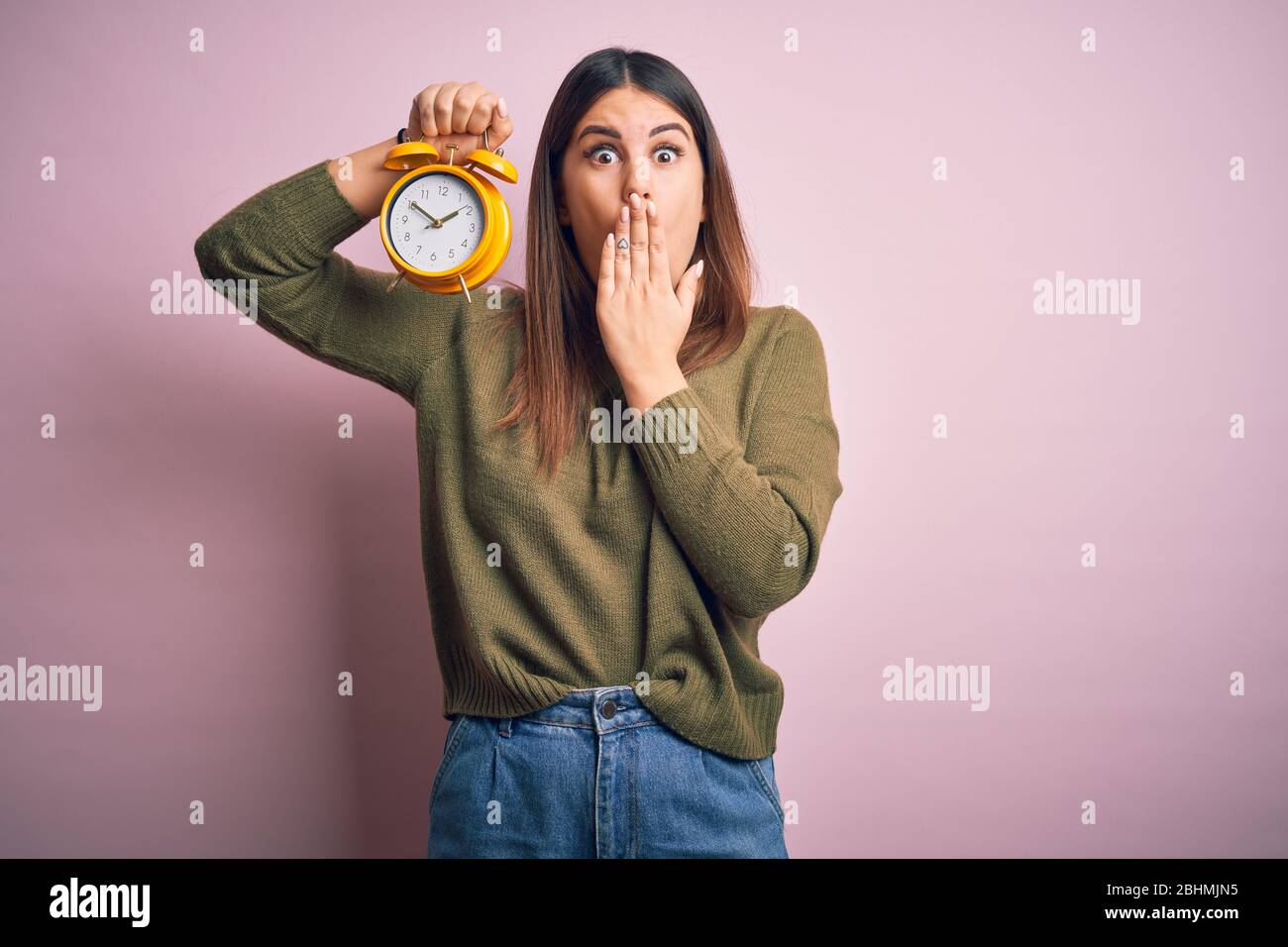 Young beautiful woman holding alarm clock standing over isolated pink ...