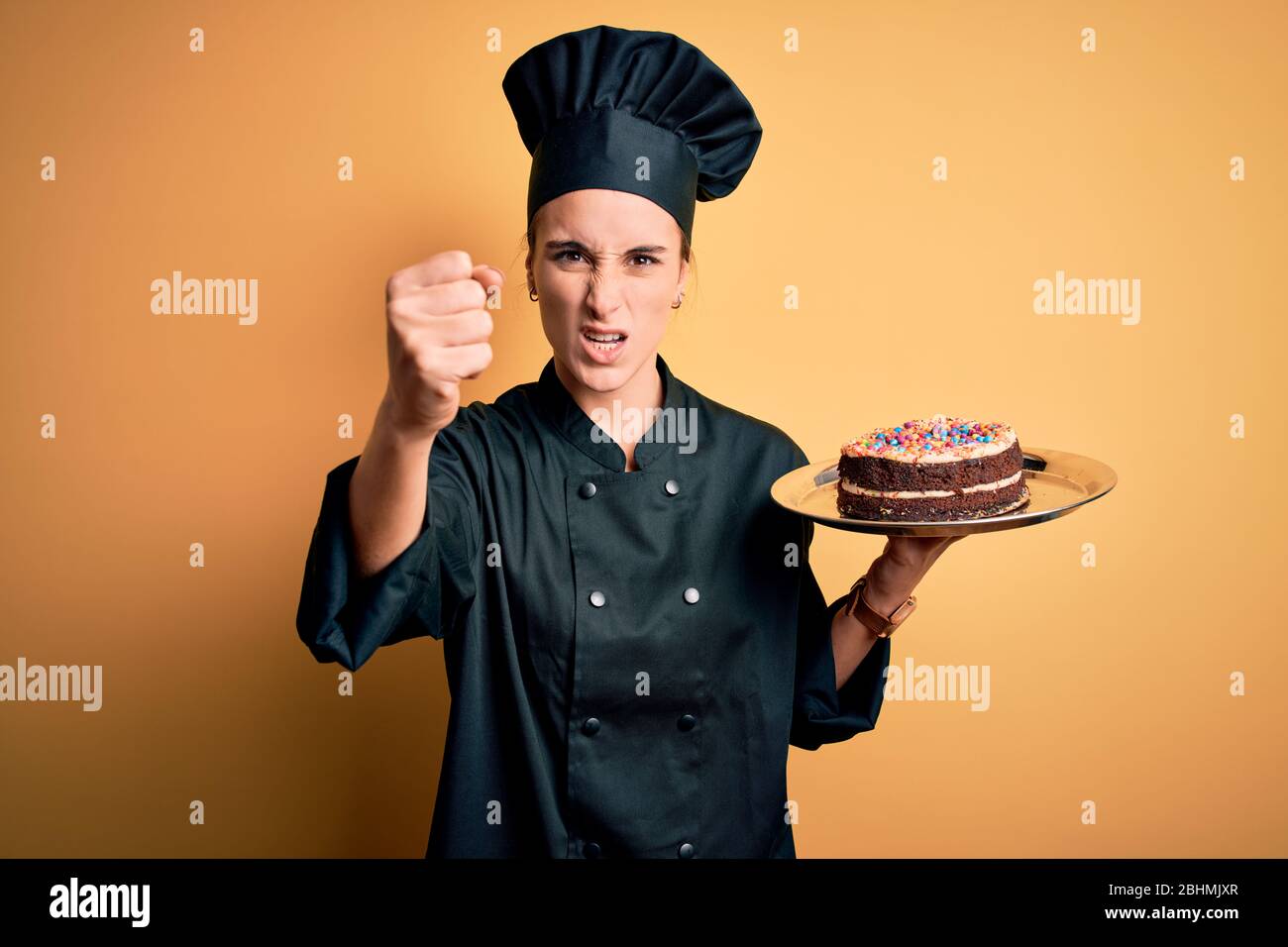 Young beautiful baker woman wearing cooker uniform and hat holding tray ...