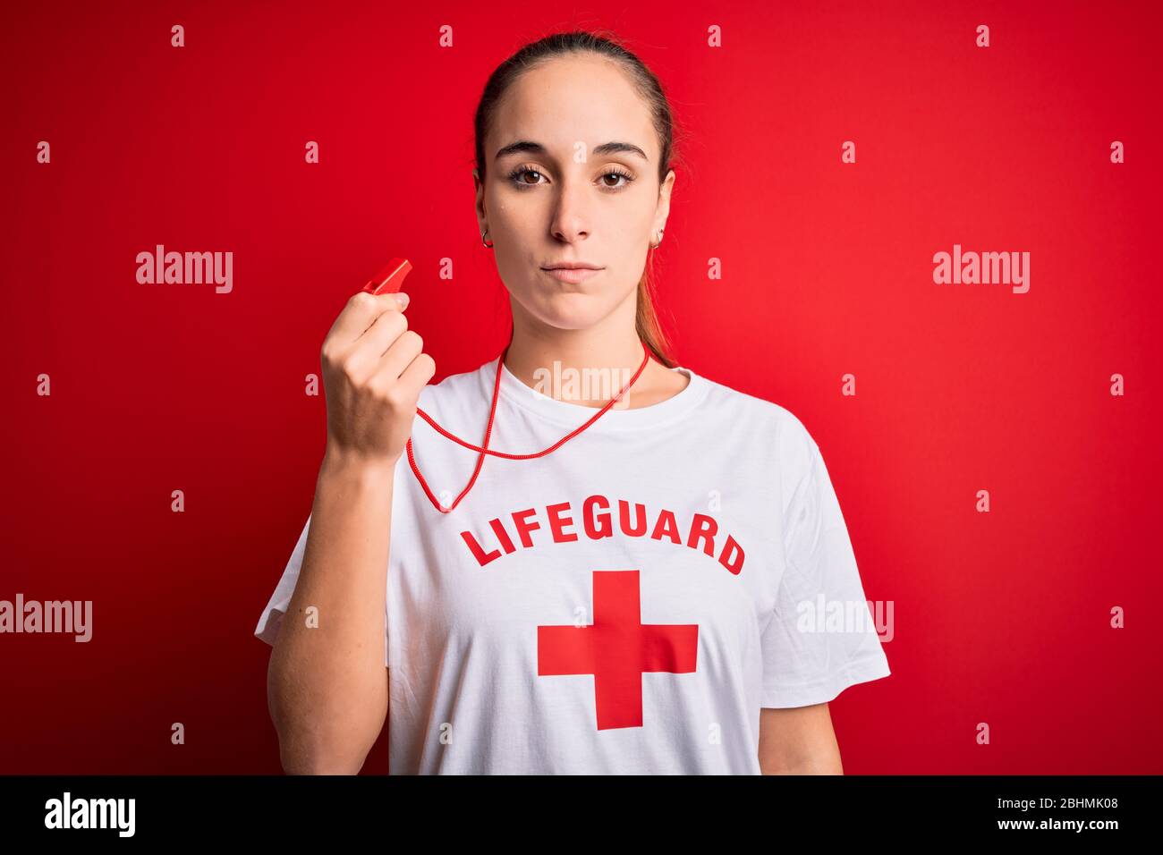 Beautiful lifeguard woman wearing t-shirt with red cross using whistle ...