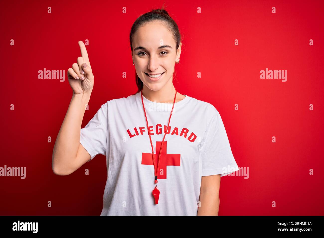 Beautiful lifeguard woman wearing t-shirt with red cross using whistle ...