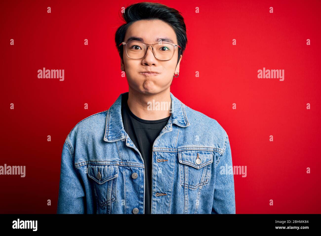 Young handsome chinese man wearing denim jacket and glasses over red ...