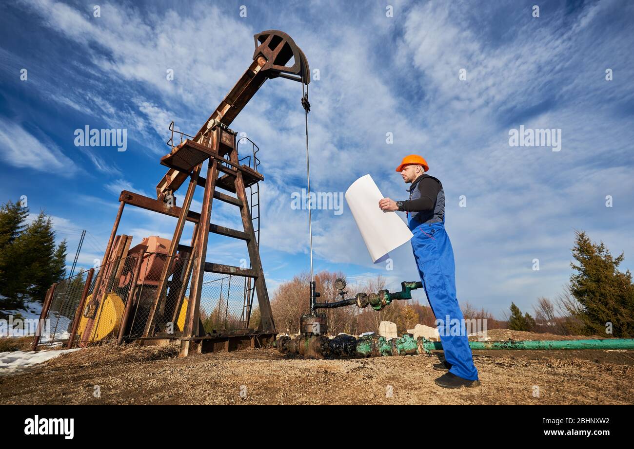 Petroleum worker holding plan of oil field and looking at oil pump jack ...