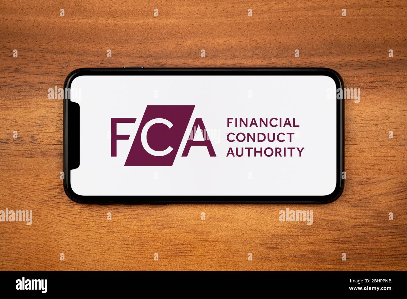 A smartphone showing the FCA logo rests on a plain wooden table ...
