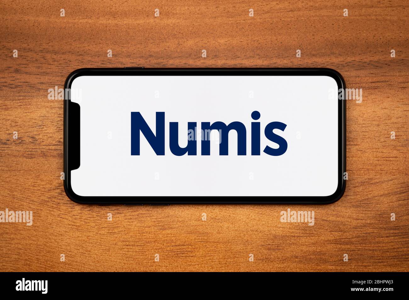 A smartphone showing the Numis logo rests on a plain wooden table ...
