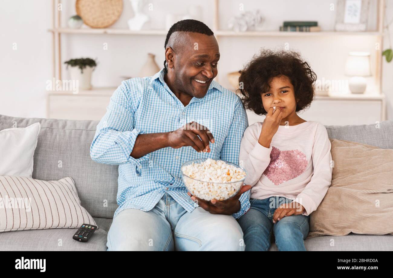 Family movie night. African American girl watching TV with her grandfather and eating popcorn at ...