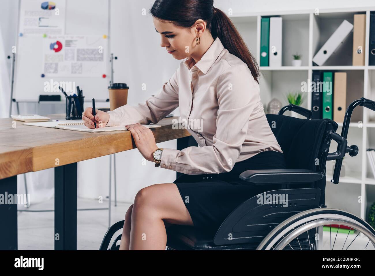 Disabled employee on wheelchair writing in notebook at table in office ...