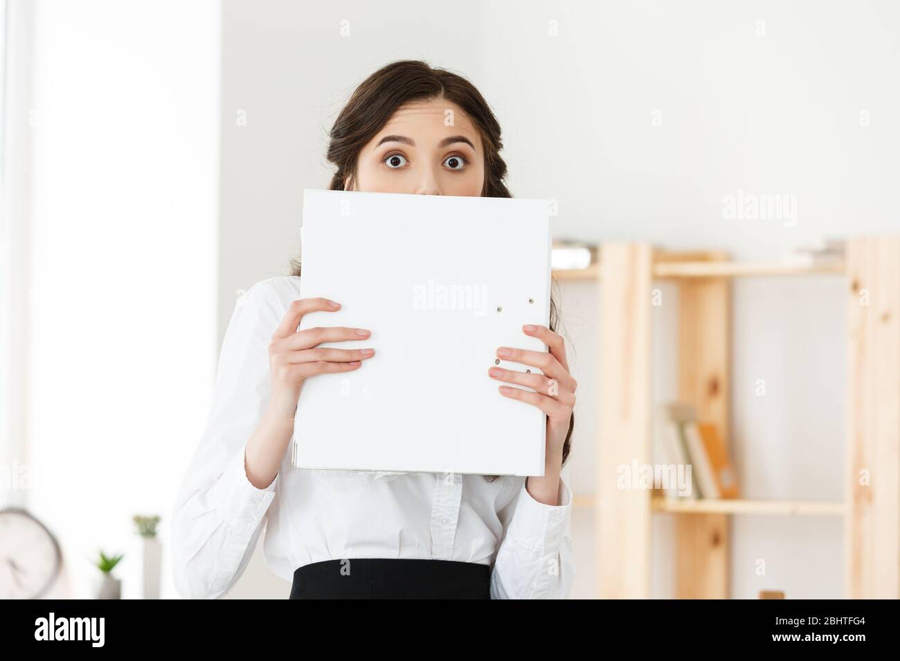 Young woman with surprised eyes peeking out from behind paper poster ...