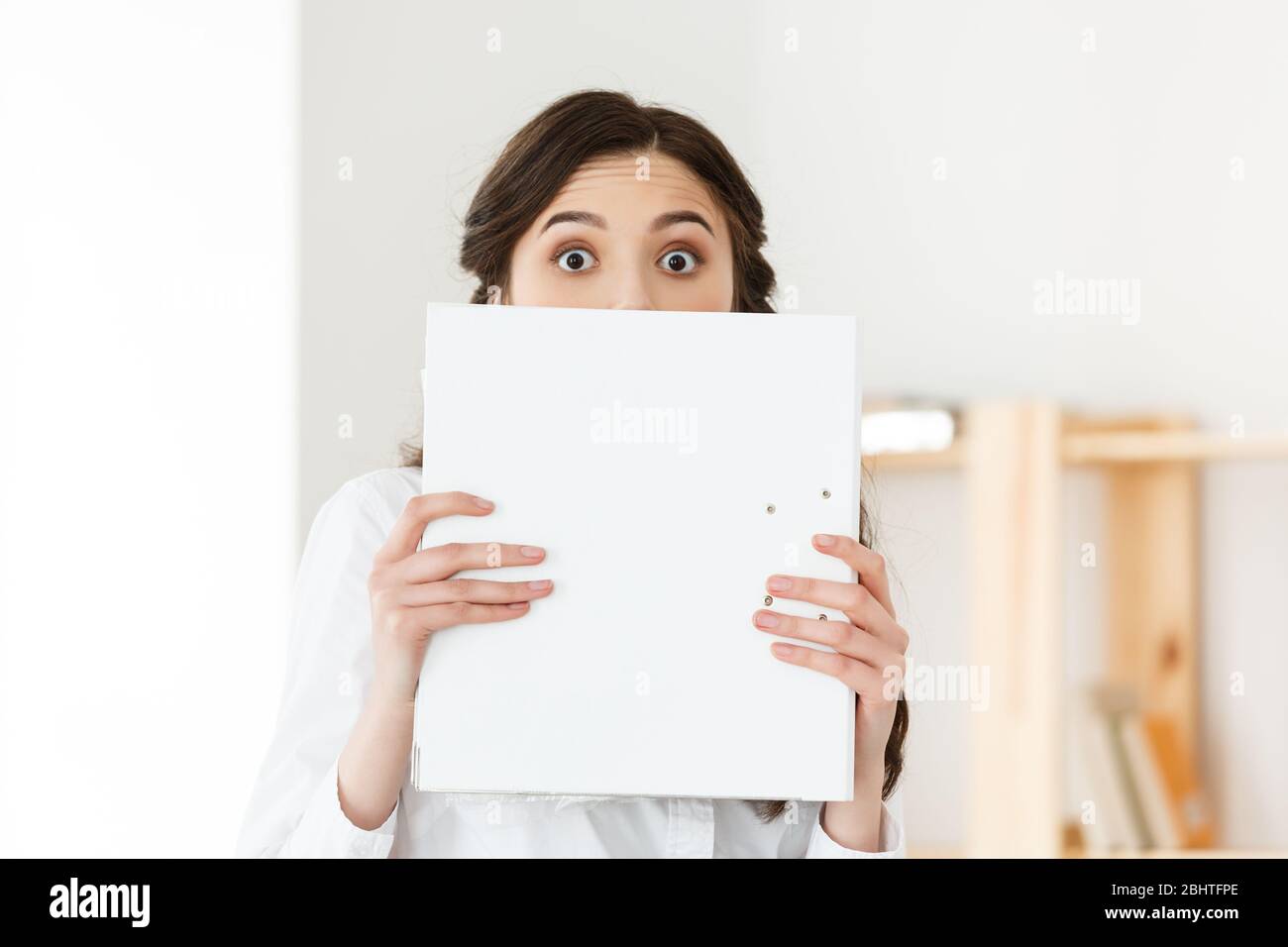 Young woman with surprised eyes peeking out from behind paper poster ...