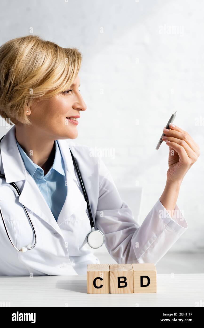 happy and mature doctor holding rolled joint near cubes with cbd ...