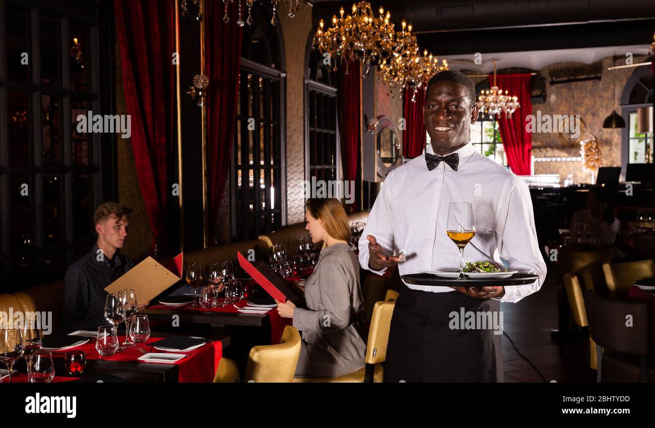 Polite waiter holding tray in restaurant with customers behind him ...
