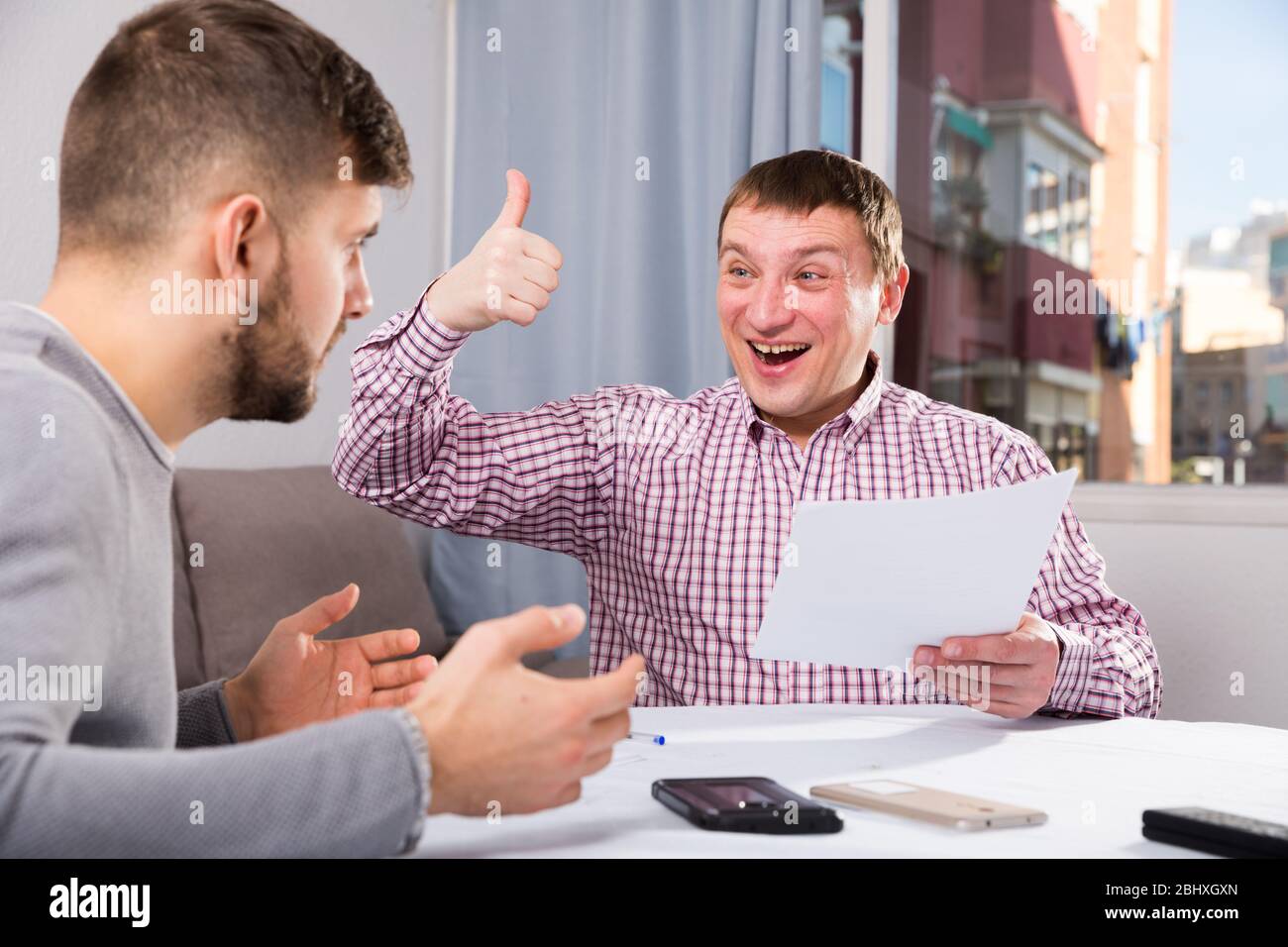 Satisfied man with male partner working with documents sitting at table ...