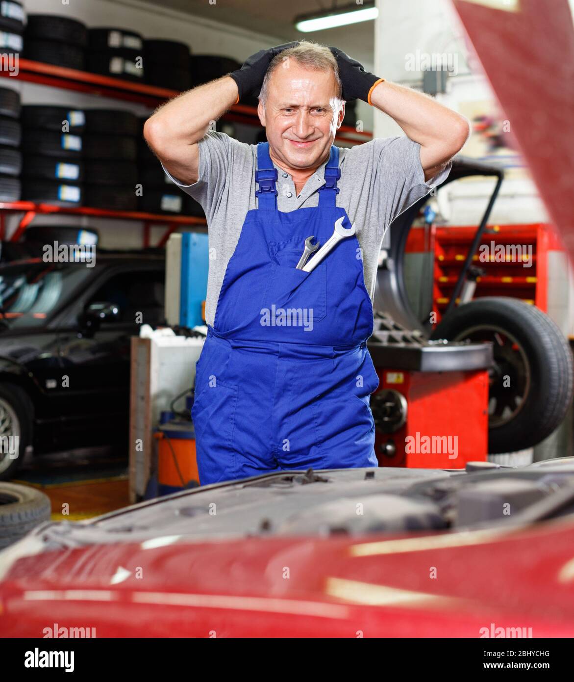 Puzzled cheerful smiling male mechanic inspecting broken car and ...