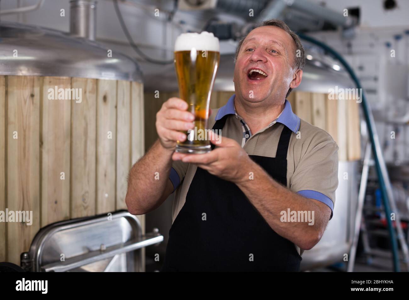 Brewer is standing with beer with foam in glass on his workplace indoor ...