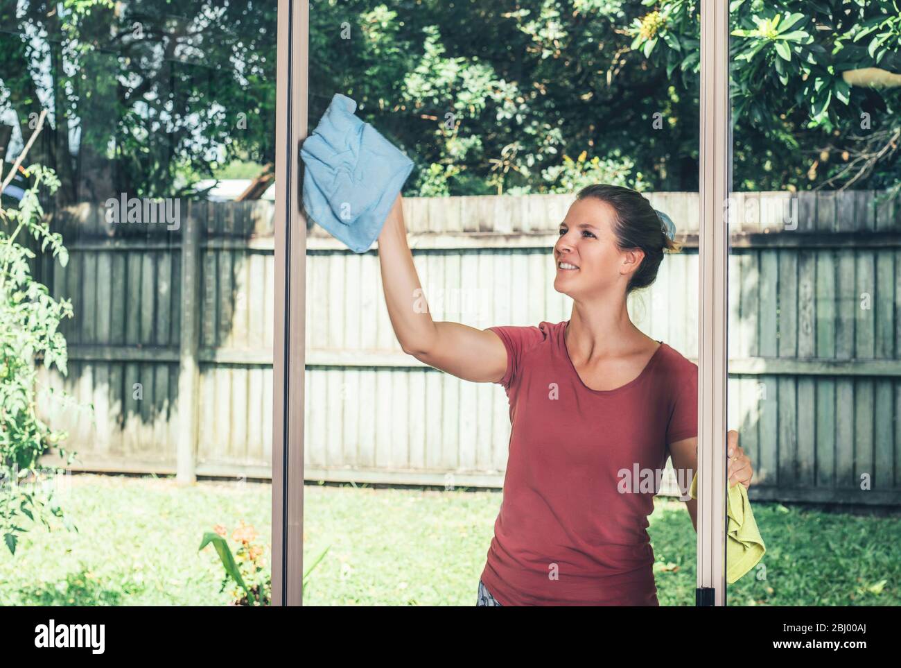 Young smiling woman is cleaning windows staying in the backyard, doing ...