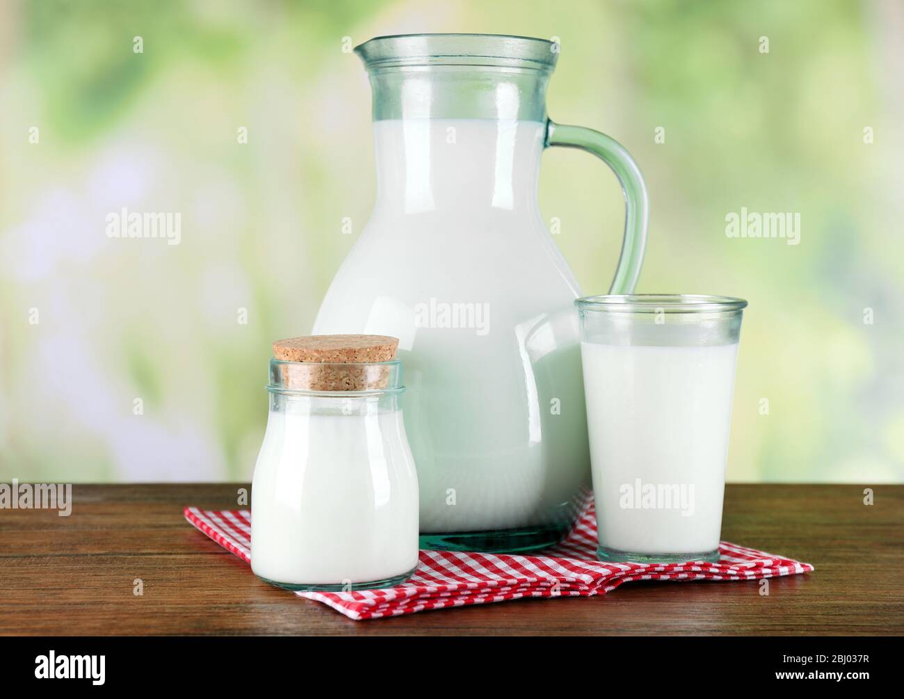 Pitcher, jar and glass of milk on wooden table, on nature background ...