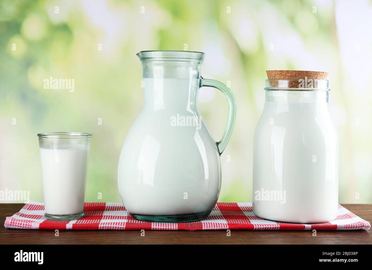 Pitcher, jar and glass of milk on wooden table, on nature background ...