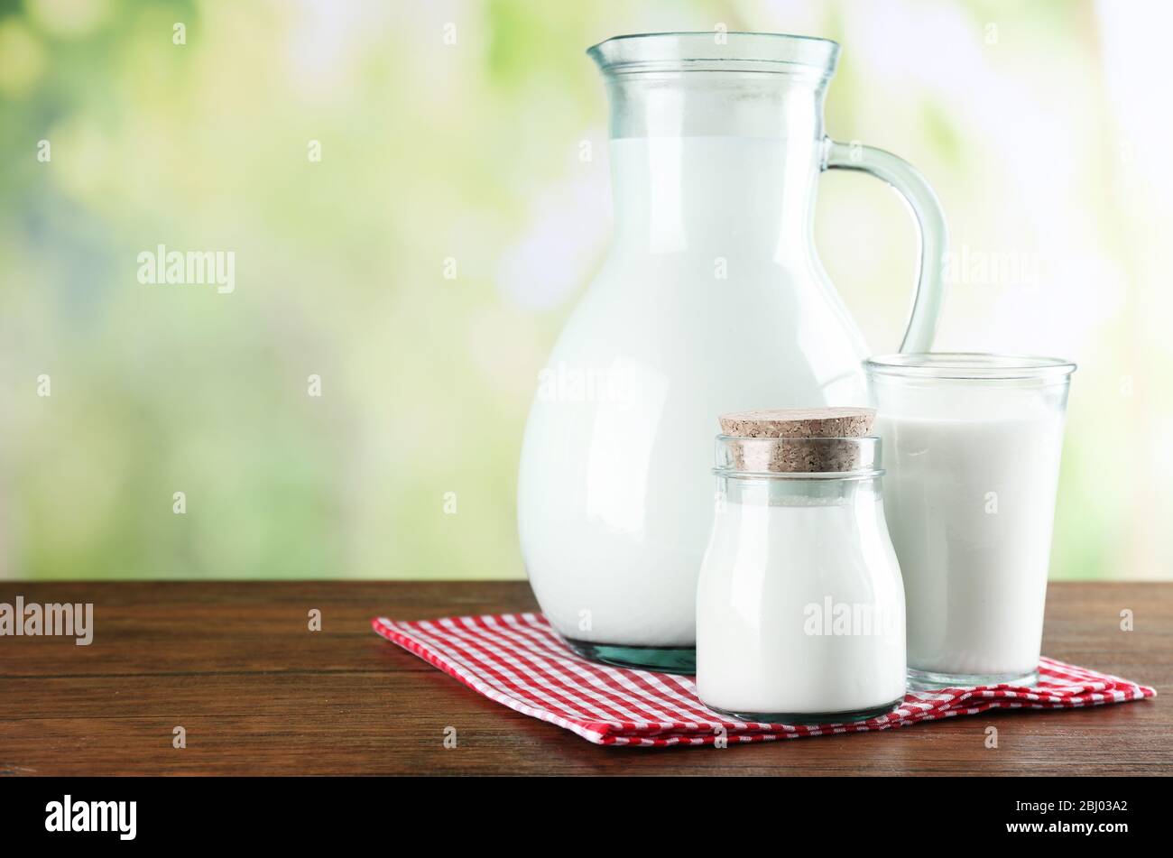 Pitcher, jar and glass of milk on wooden table, on nature background ...