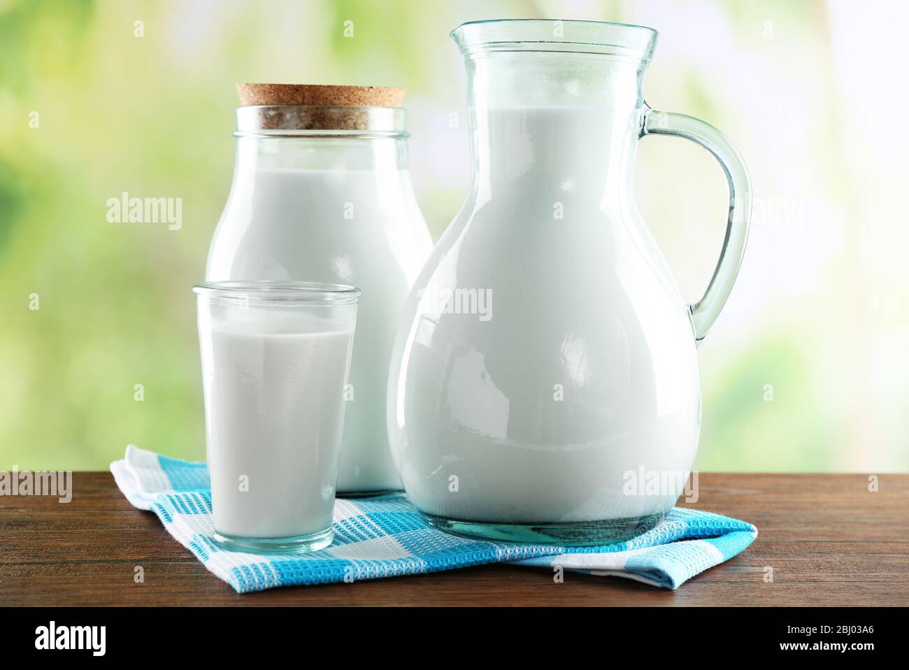 Pitcher, jar and glass of milk on wooden table, on nature background ...