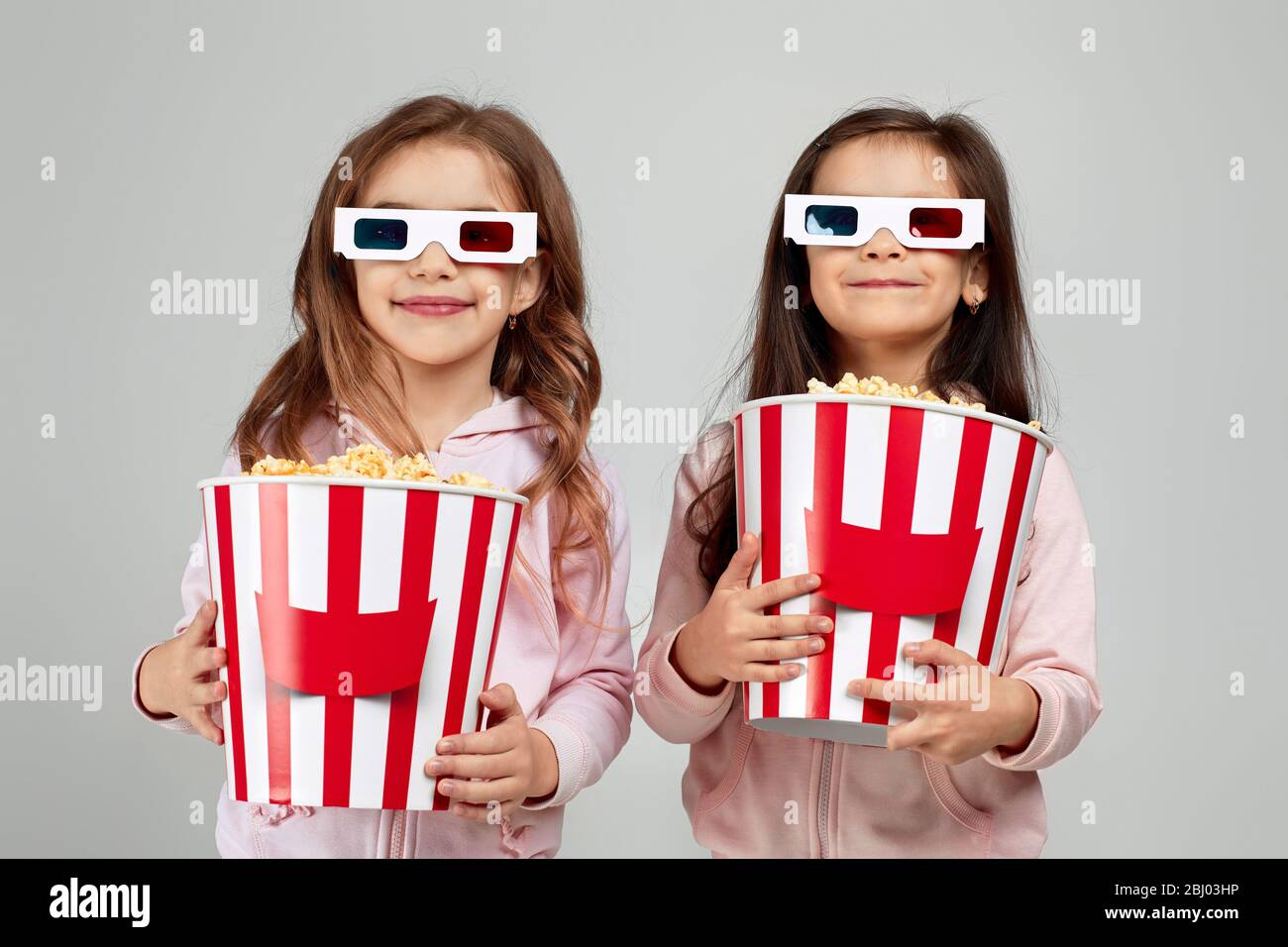 two beautiful caucasian smiling little girls in red-blue 3d glasses holding popcorn buckets and ...