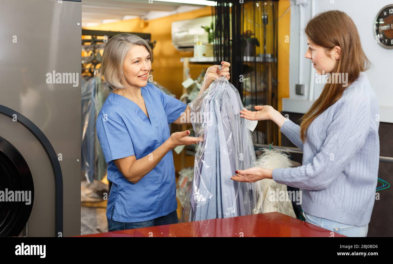 Adult worker of dry-cleaning salon giving woman her clean clothes in ...