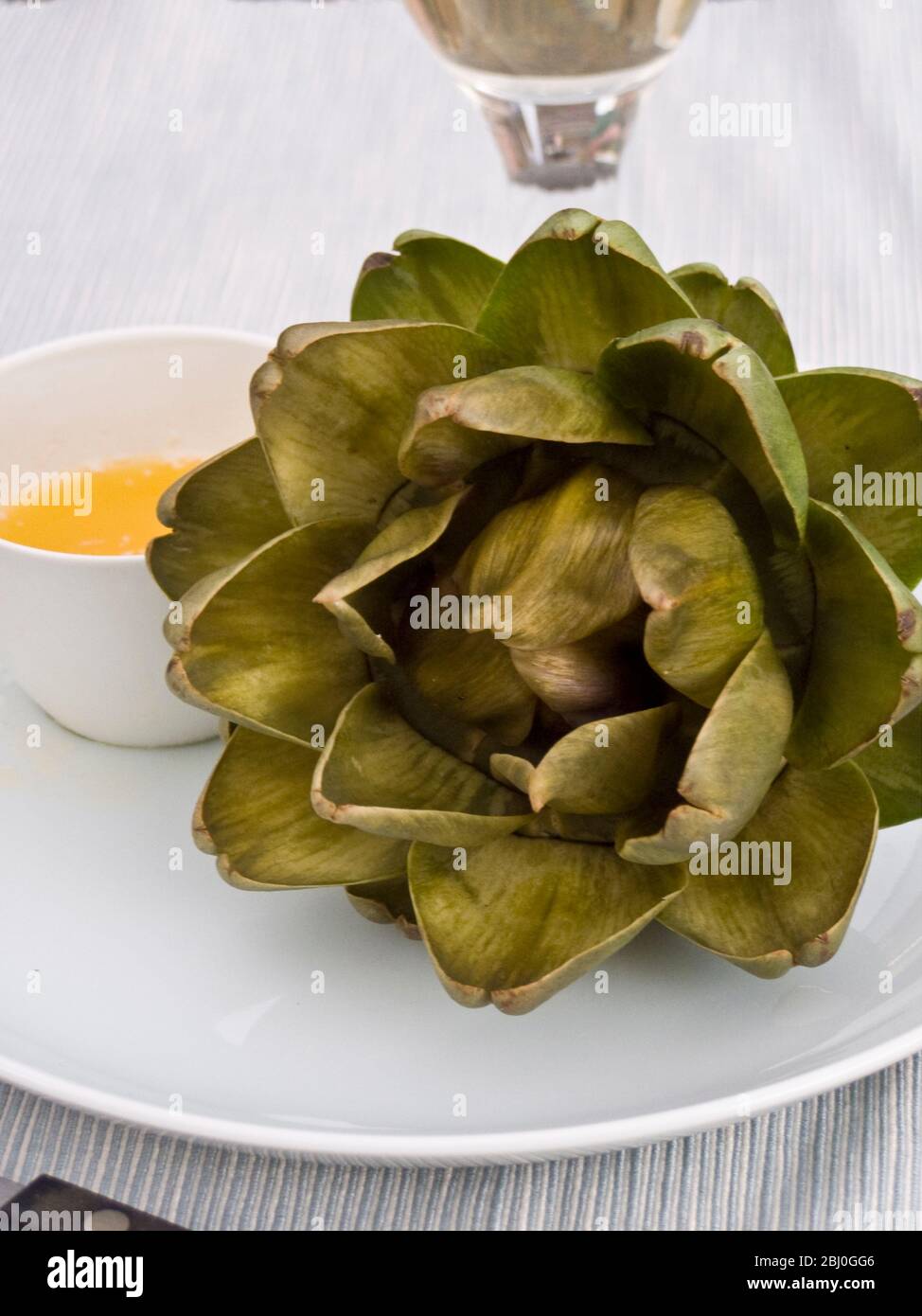 Whole cooked globe artichoke served on white plate with pot of melted ...
