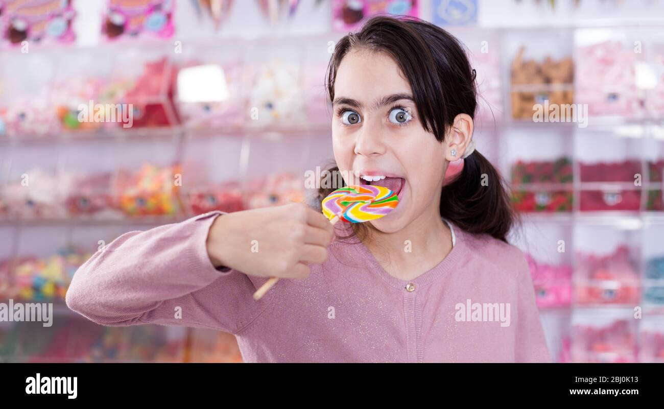 Positive small girl holding sweet colored candy on stick in candy shop ...