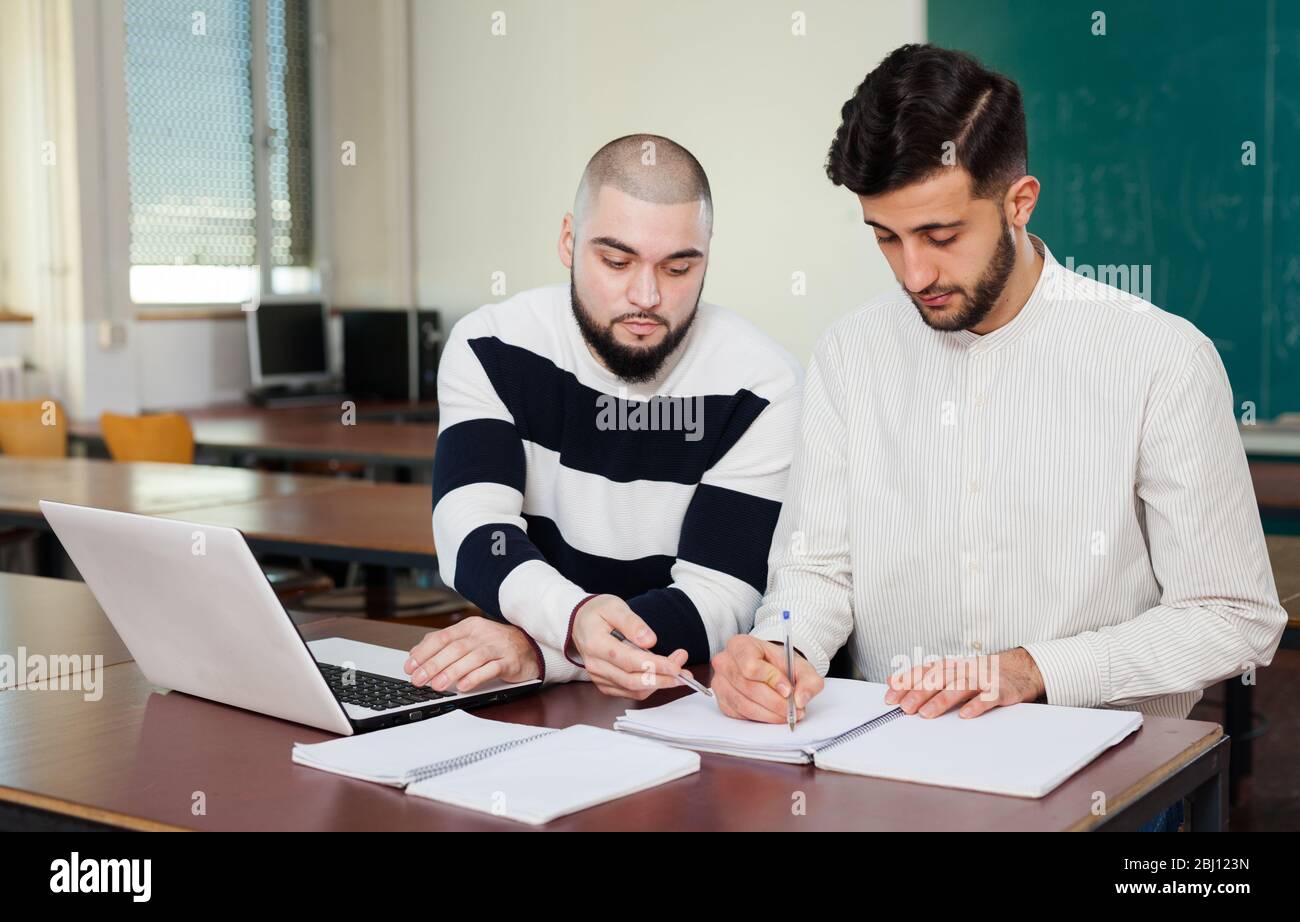 Two young handsome students working with laptop at desk in classroom ...