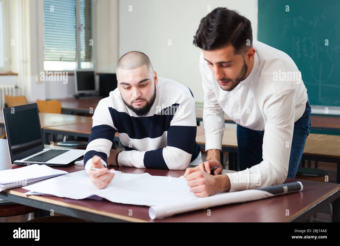 Two young handsome students working with laptop at desk in classroom ...