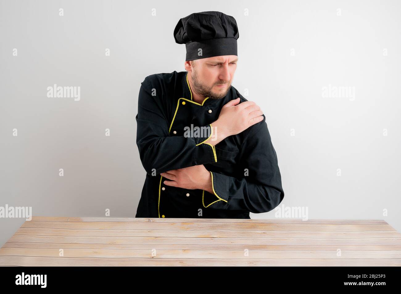 Young male chef in black uniform has shoulder pain posing on a white ...