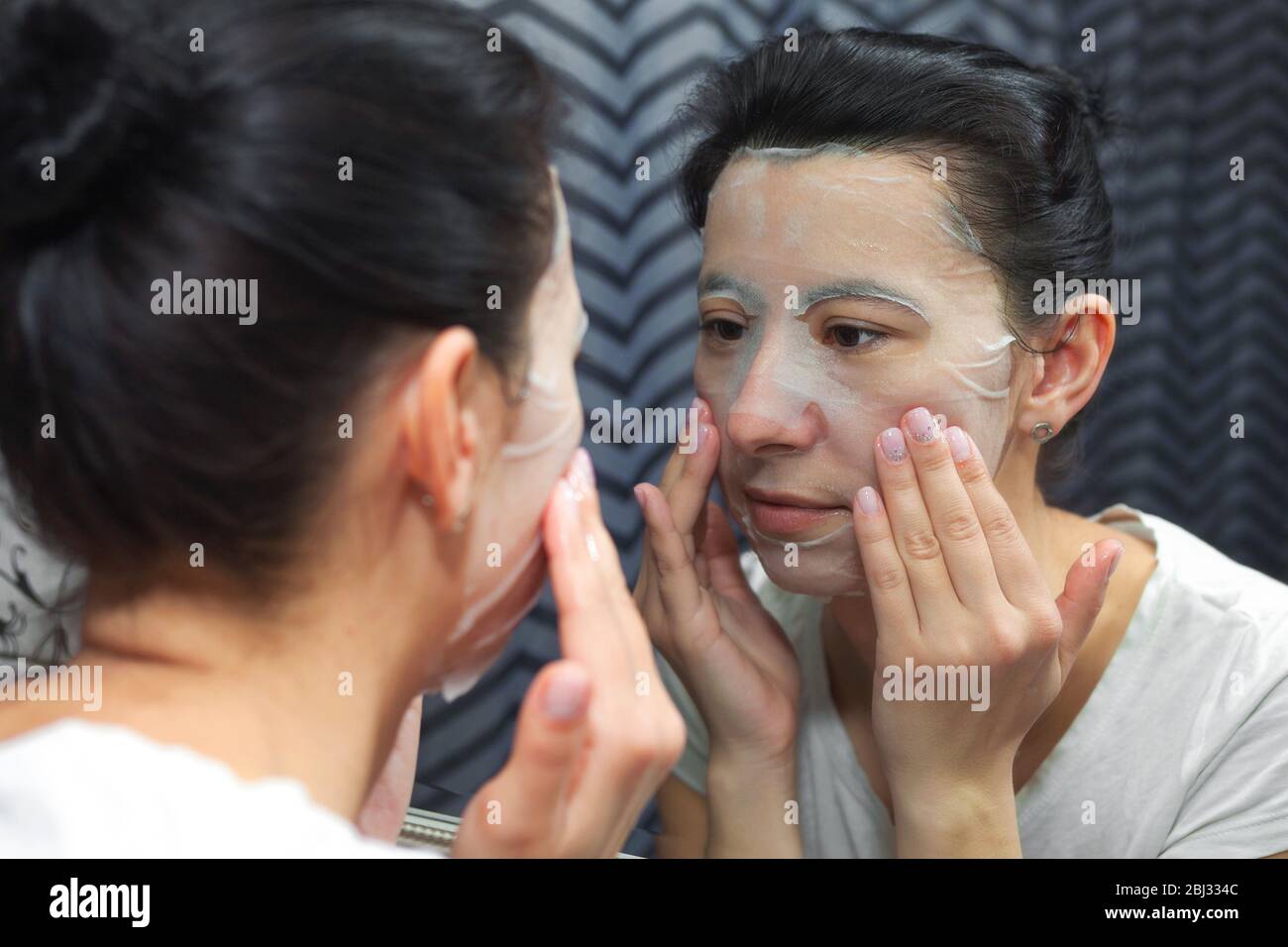 A girl in a cosmetic mask looks in the mirror at her reflection. Makes ...
