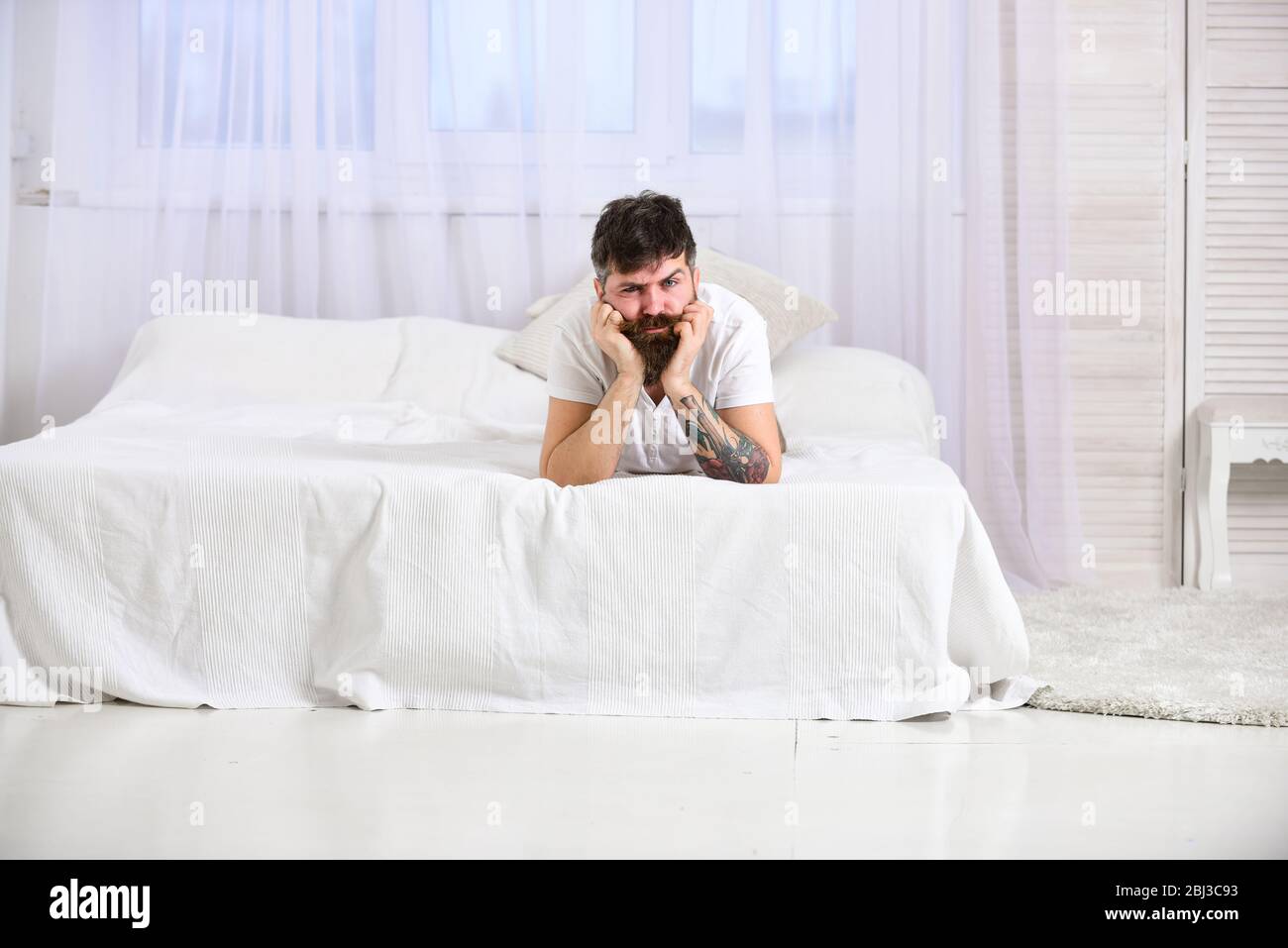 Guy on sleepy face laying on edge of bed on white sheets. Bedroom and ...