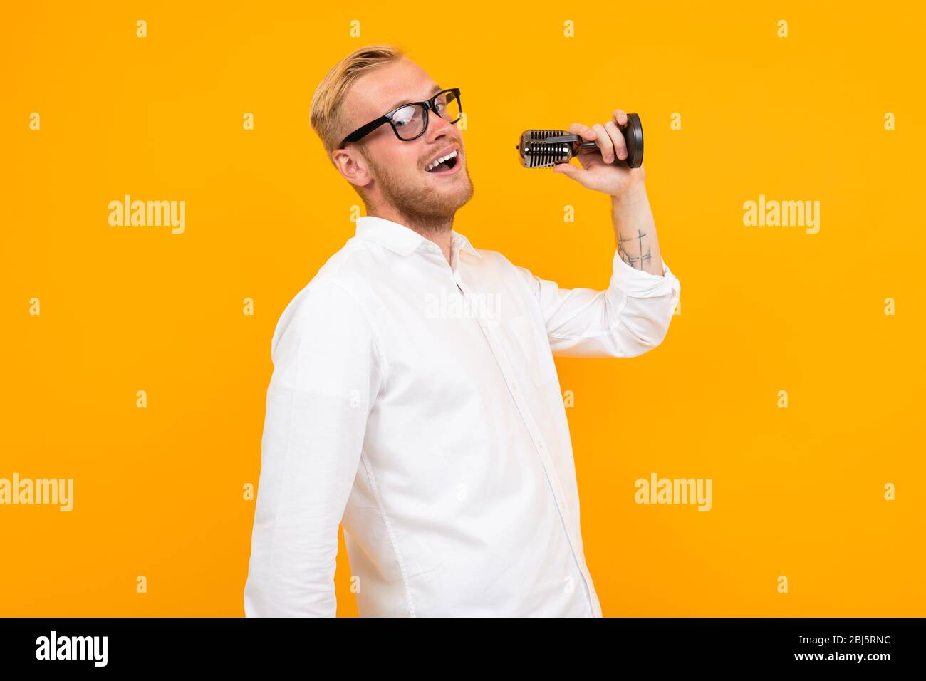 blond guy in a white shirt sings into a retro microphone on a yellow ...