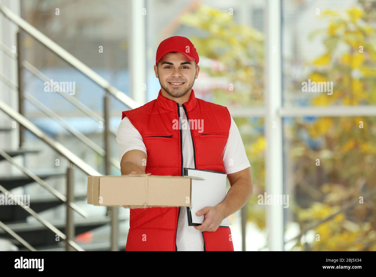 Delivery concept - postman in red uniform holding package near stairs ...