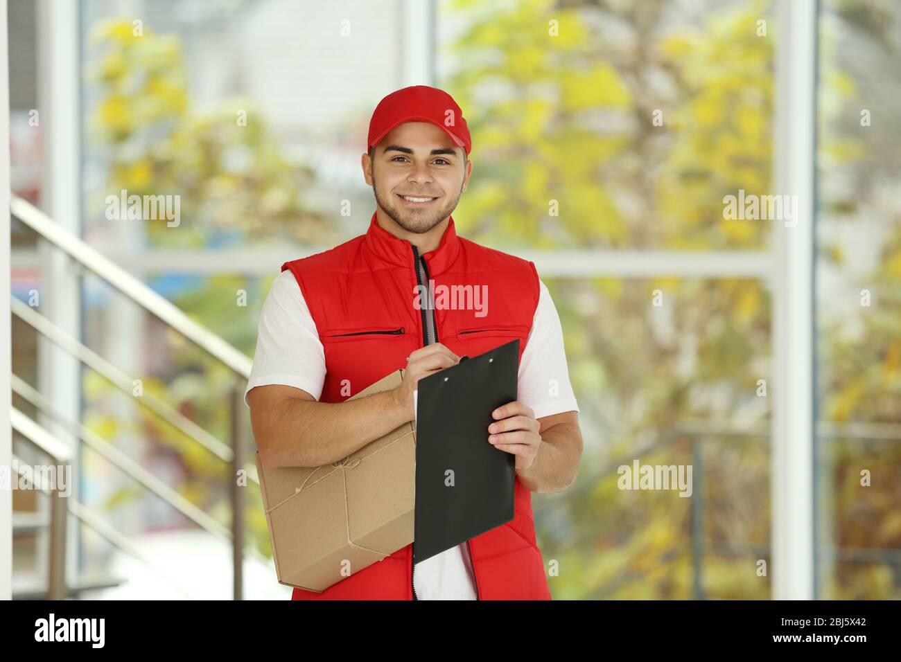 Delivery concept - postman in red uniform holding package near stairs ...