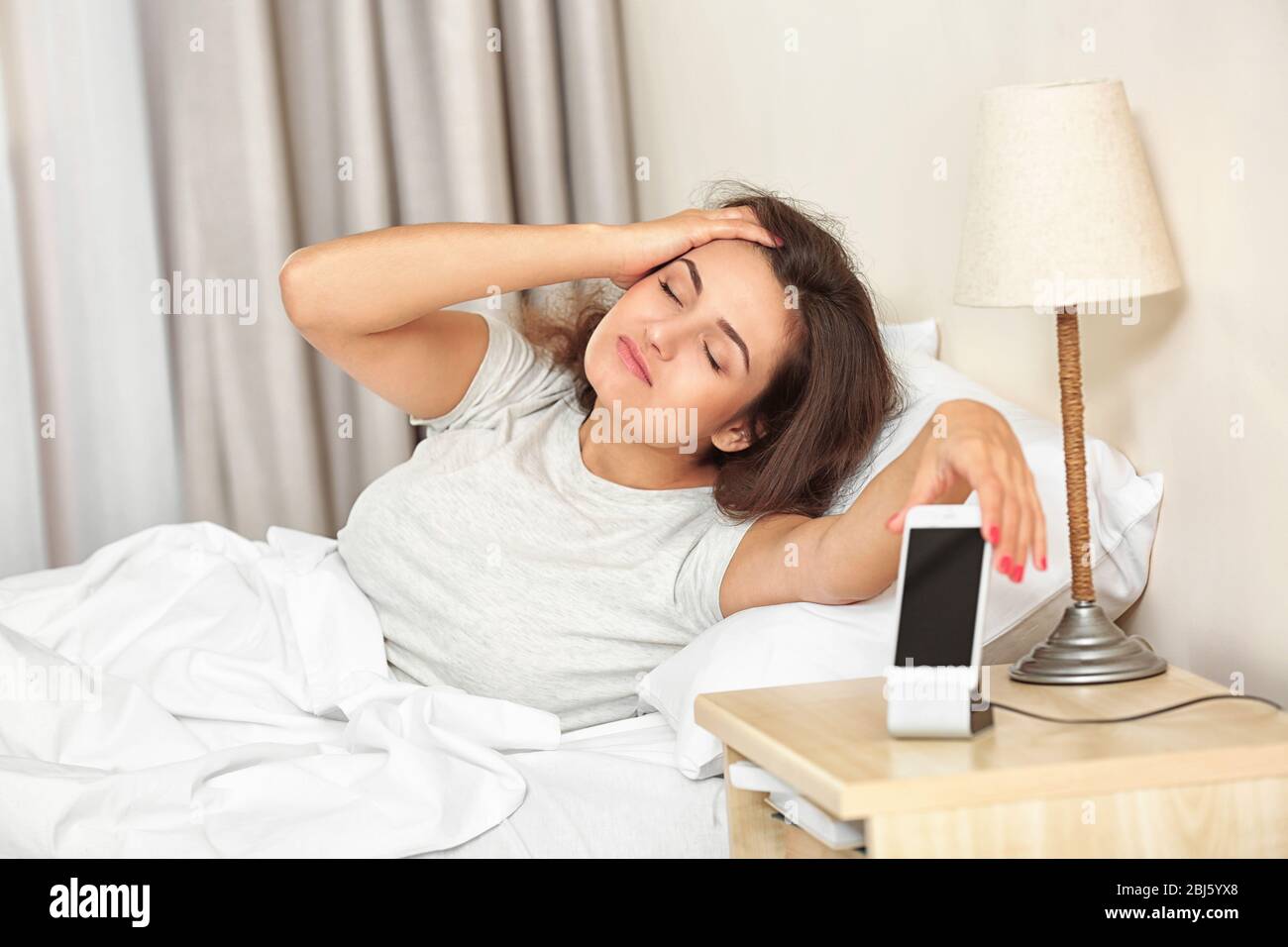 Beautiful young woman waking up with mobile alarm clock Stock Photo - Alamy