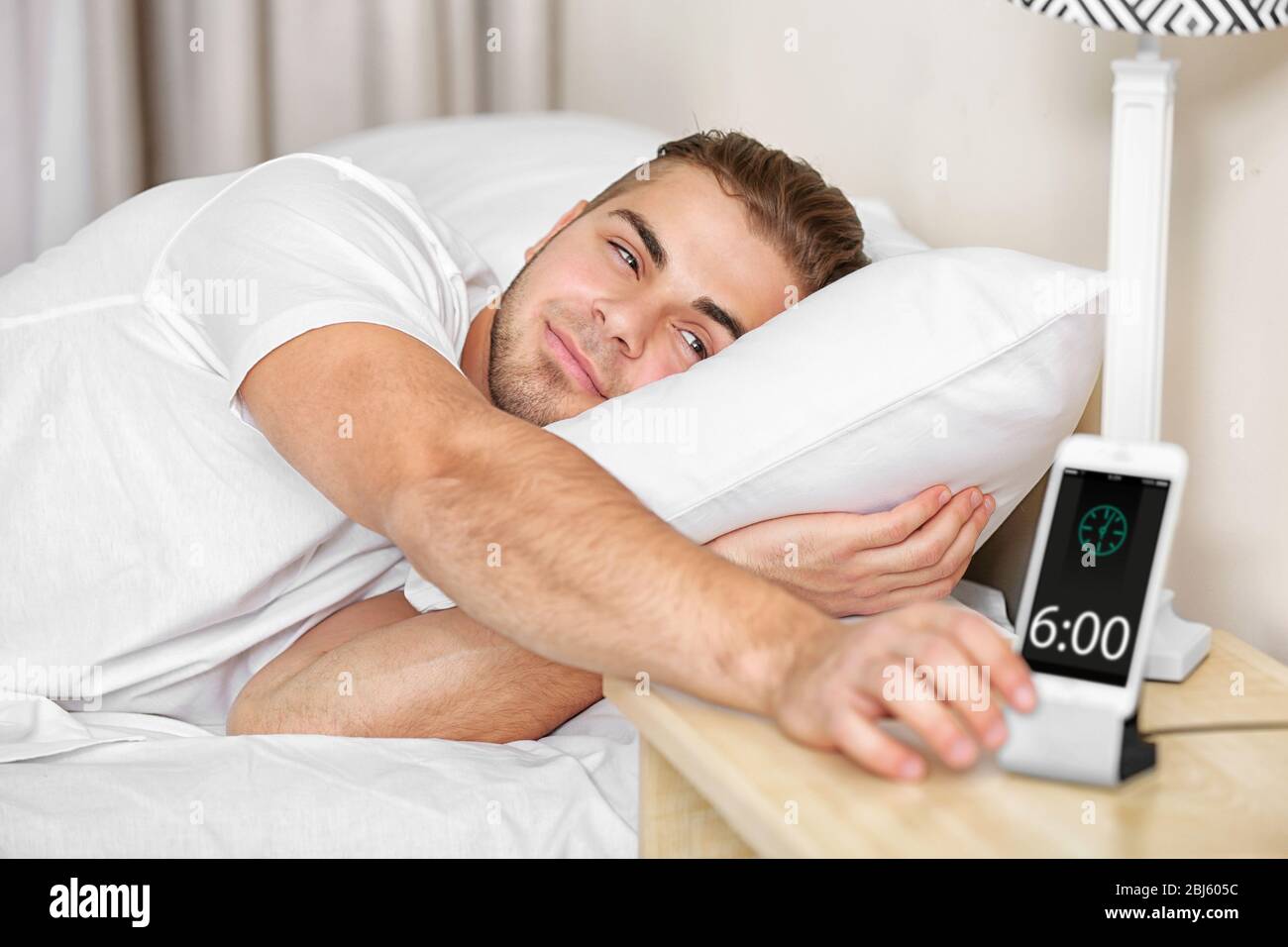 Beautiful young man waking up with mobile alarm clock Stock Photo - Alamy
