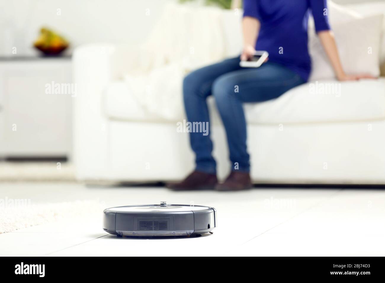 Robotic vacuum cleaner cleaning the room while woman sitting on sofa ...