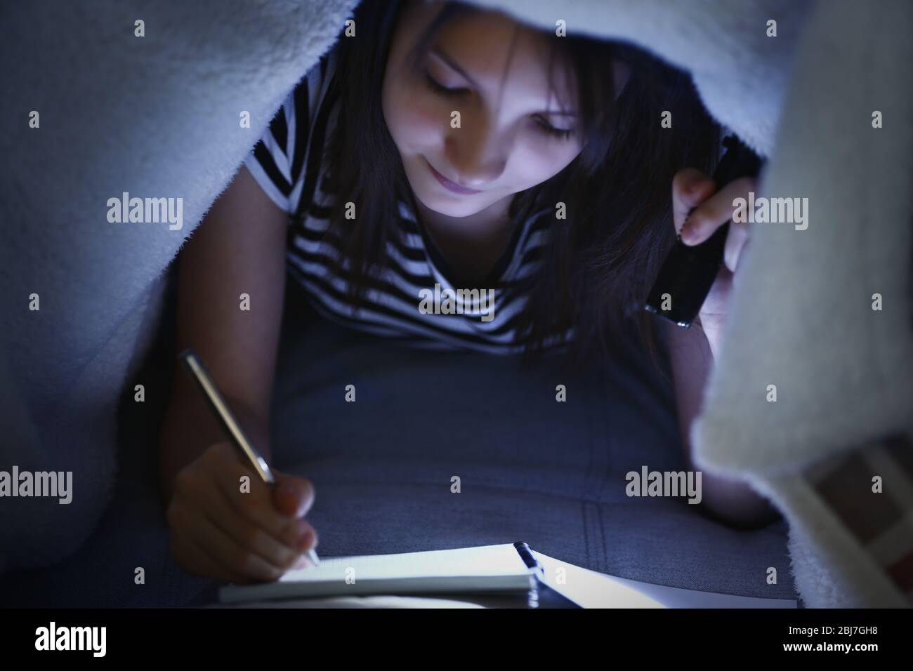 Beautiful girl writing in notebook with flashlight under the blanket ...