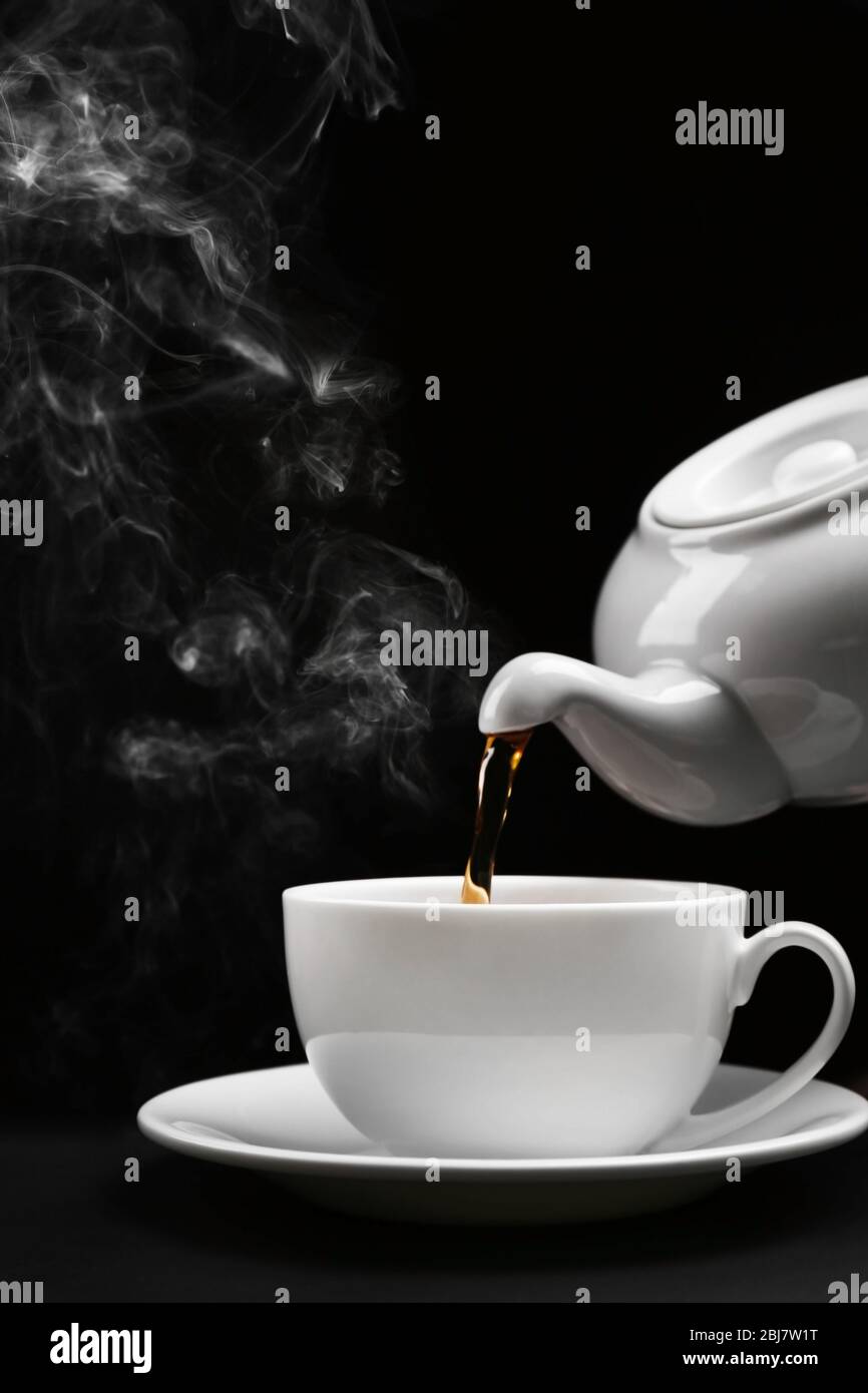 Pouring hot tea from a kettle into a cup on black background, close up ...