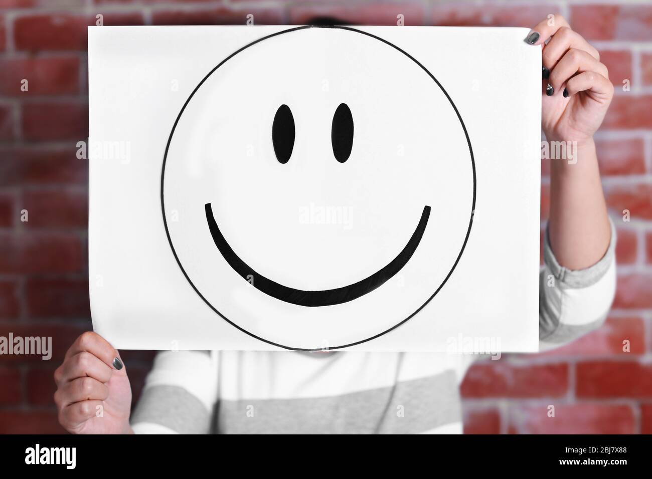 Woman showing a happy emoticon in front of face against a brick wall ...