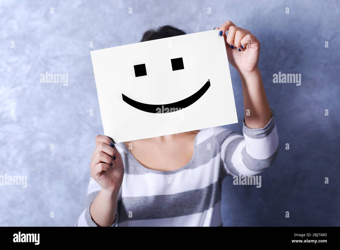 Woman showing a happy emoticon in front of face against a grey textured ...