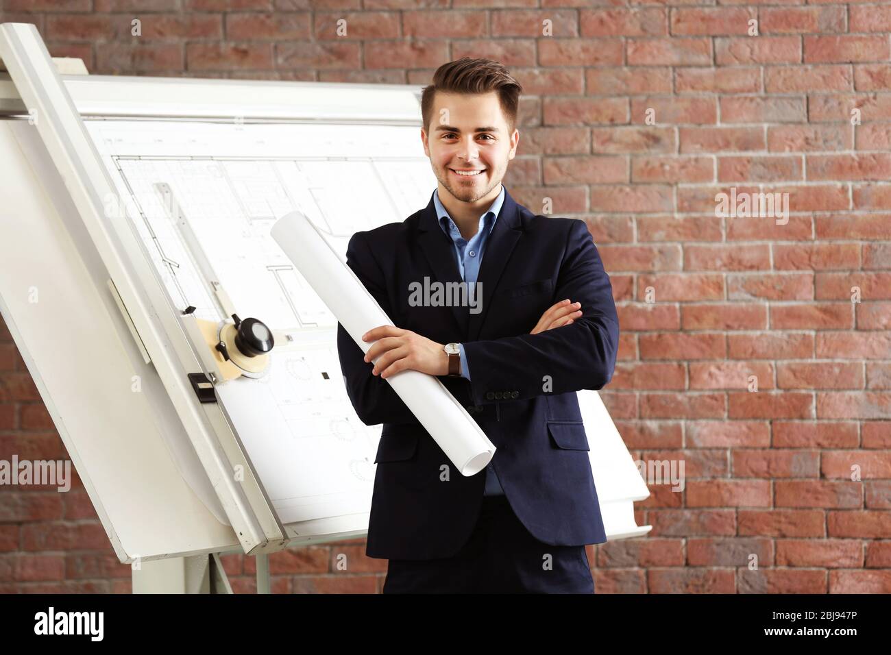 Young engineer holding blueprint near panel board on brick wall ...