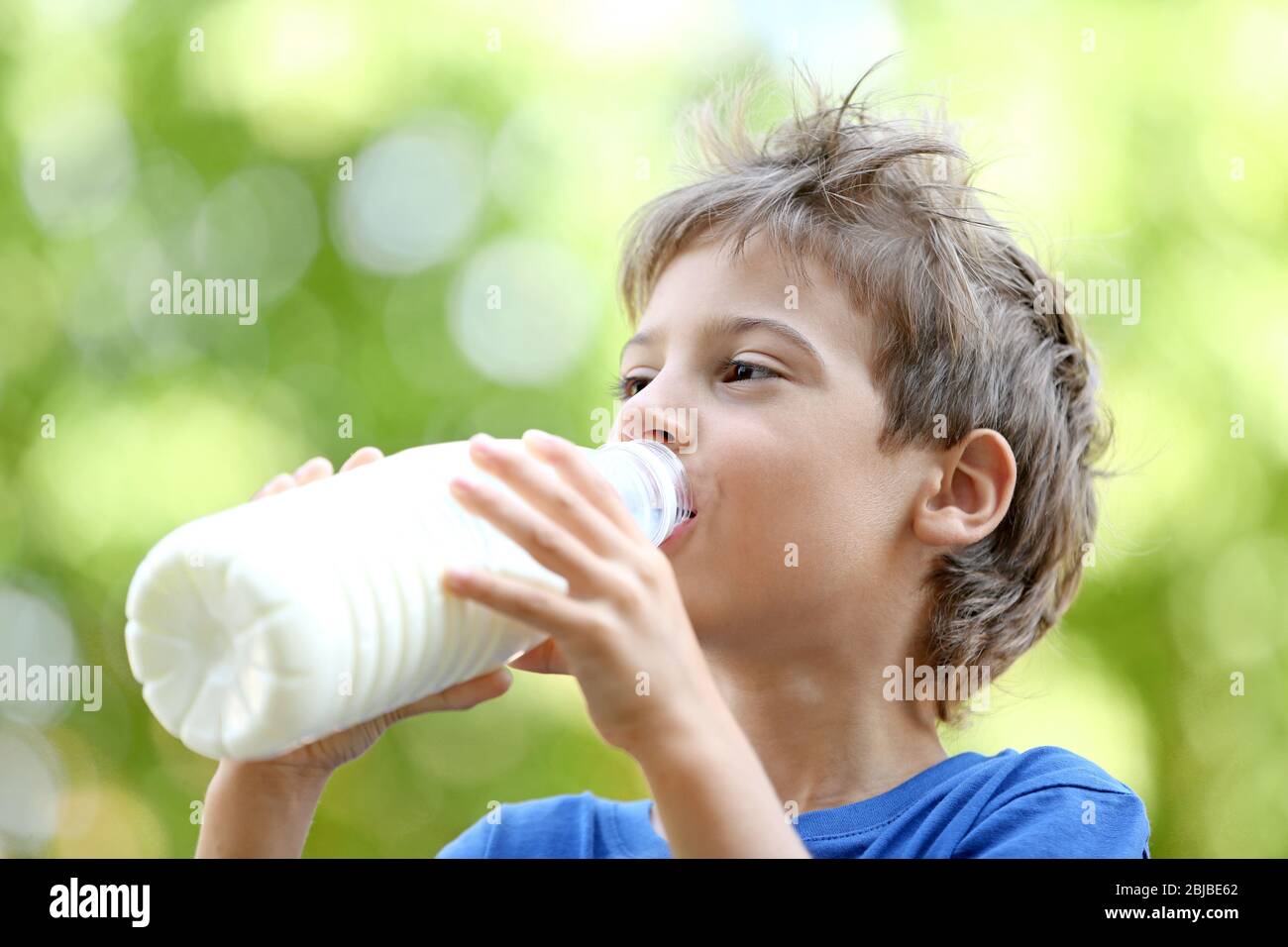 Cheerful kid in blue shirt drinking milk from plastic bottle on blurred ...