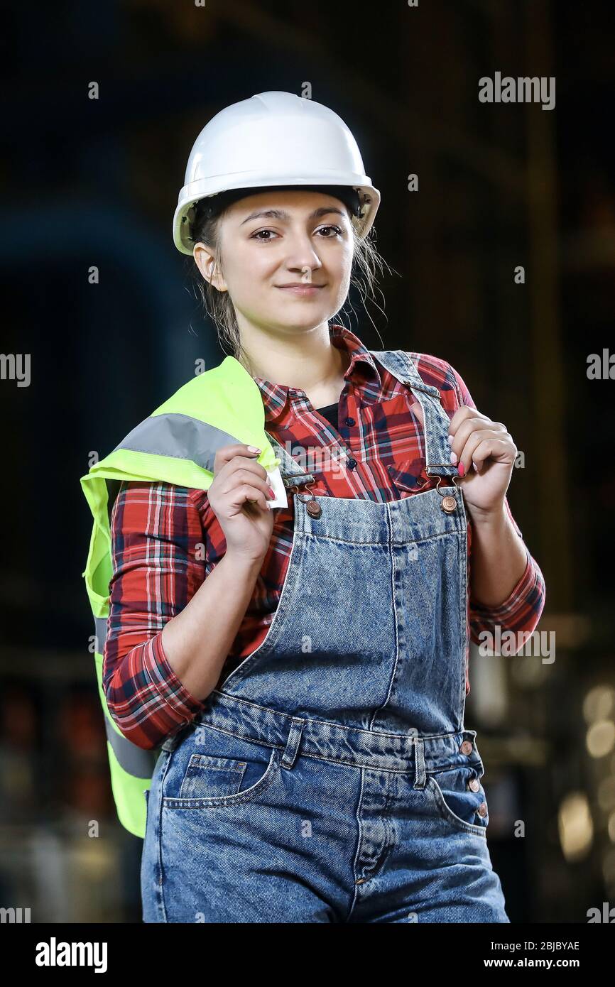 Young girl in a work dress and white hard hat in a factory. Woman in a ...